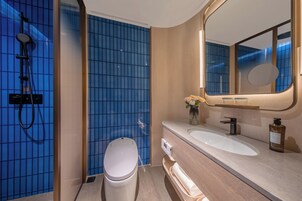 Casa de banho moderna com chuveiro em azulejo azul, sanita branca, lavatório com espelho e toucador de madeira.