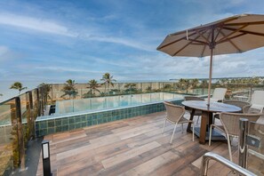 Uma área de piscina no terraço com guarda-corpo de vidro, deck de madeira e uma mesa com cadeiras.