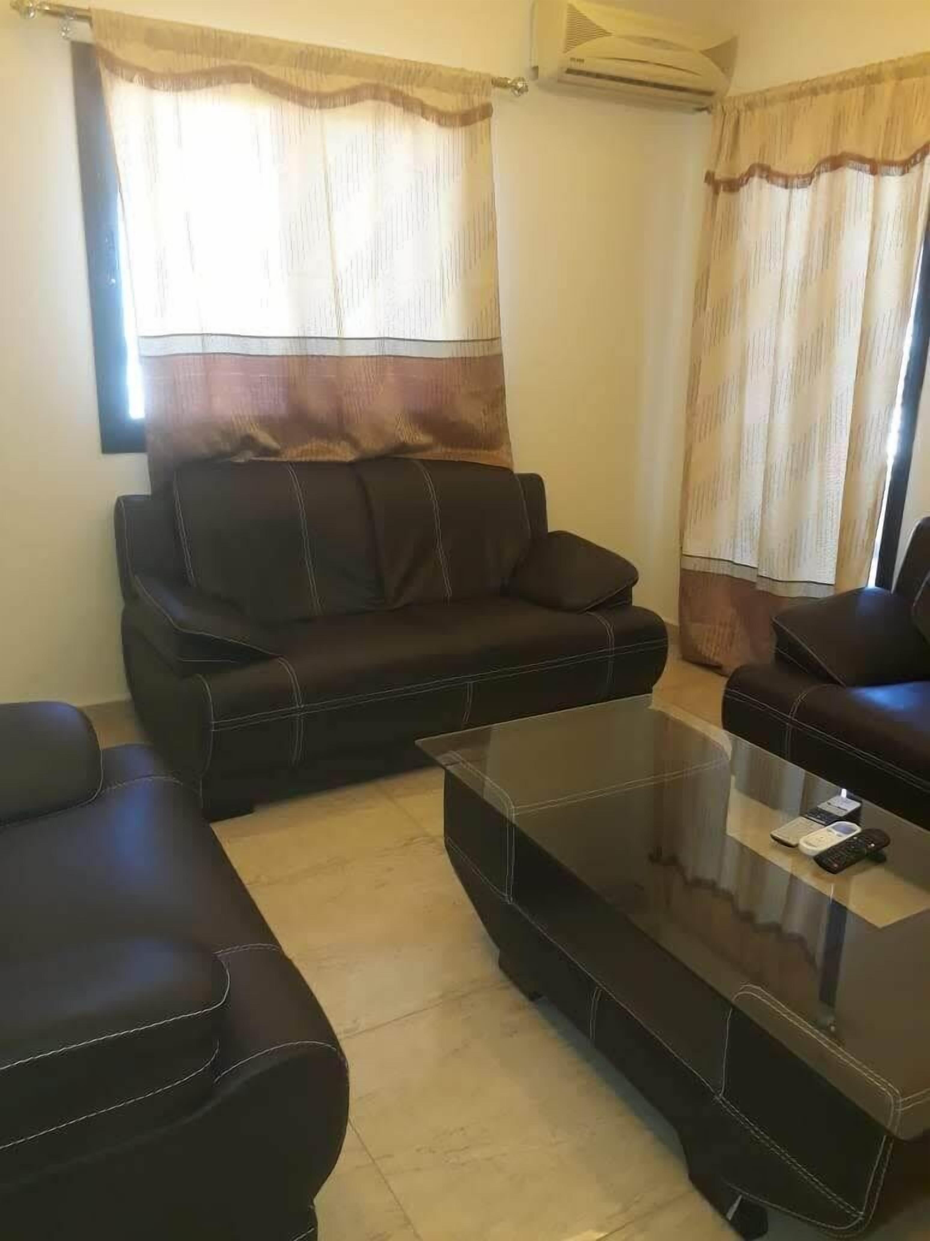 Uma sala de estar com um sofá marrom escuro, uma mesa de centro de vidro e uma mesinha lateral com um controle remoto.