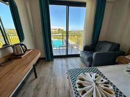 Quarto de hotel com um banco de madeira, uma mesa com uma toalha, um sofá e vista para uma piscina através de uma porta de correr.