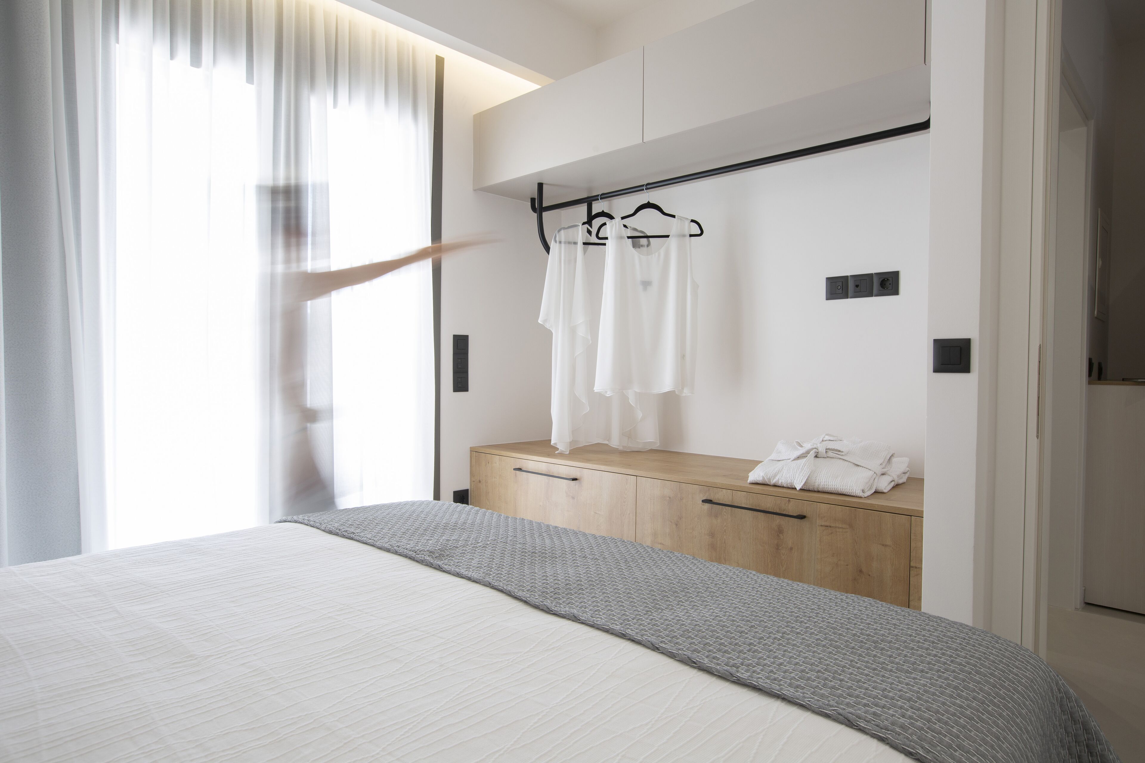 Um quarto moderno com cama, cômoda de madeira, porta-toalhas e janela com cortinas leves.