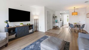 Uma sala de estar moderna com televisão de tela plana, sofás cinzas, piso de madeira e um espaço de jantar com mesa e cadeiras.