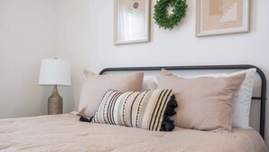 Uma cama bem arrumada com um edredom bege e duas almofadas, um abajur e duas fotos emolduradas na parede.