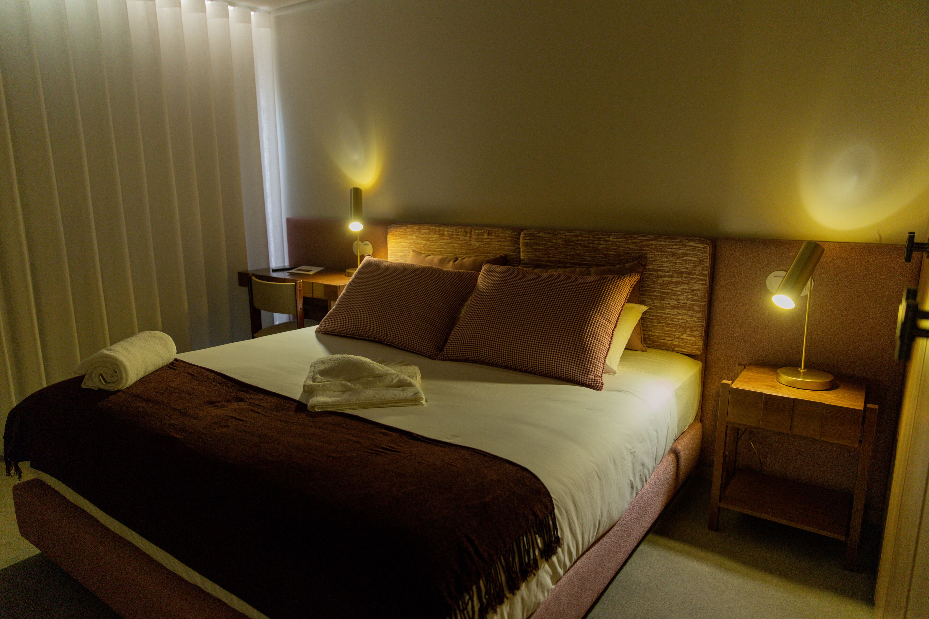 Cama bem arrumada com manta e almofadas marrons, duas luminárias de cabeceira e uma janela com persianas.