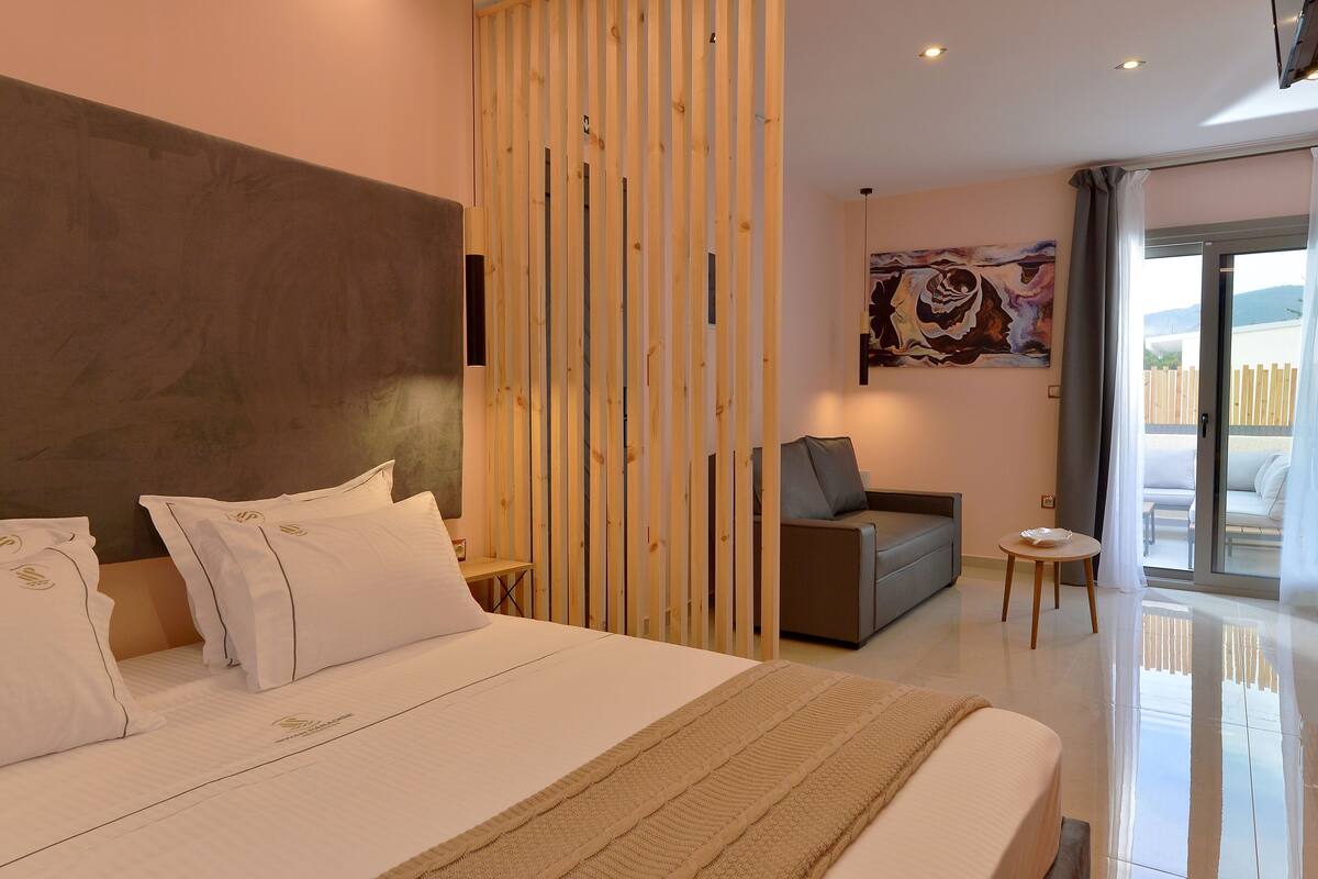Quarto de hotel moderno com uma cama grande, um sofá, uma mesa pequena e vista através de uma porta de vidro.