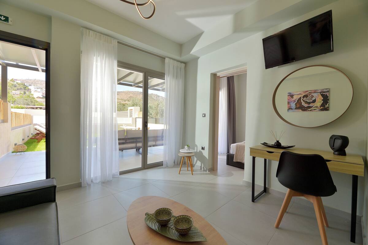 Quarto de hotel moderno com uma grande porta de vidro deslizante para a varanda, uma escrivaninha de madeira com uma cadeira, uma TV de tela plana na parede e uma pequena mesa redonda com objetos decorativos.