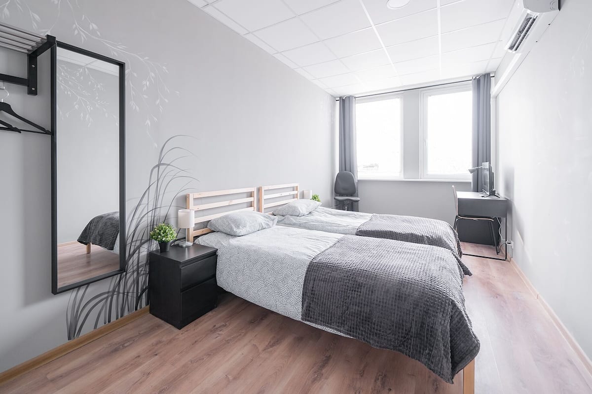 Quarto moderno com cama de madeira, mesa de cabeceira com abajur, espelho e janela com cortinas.