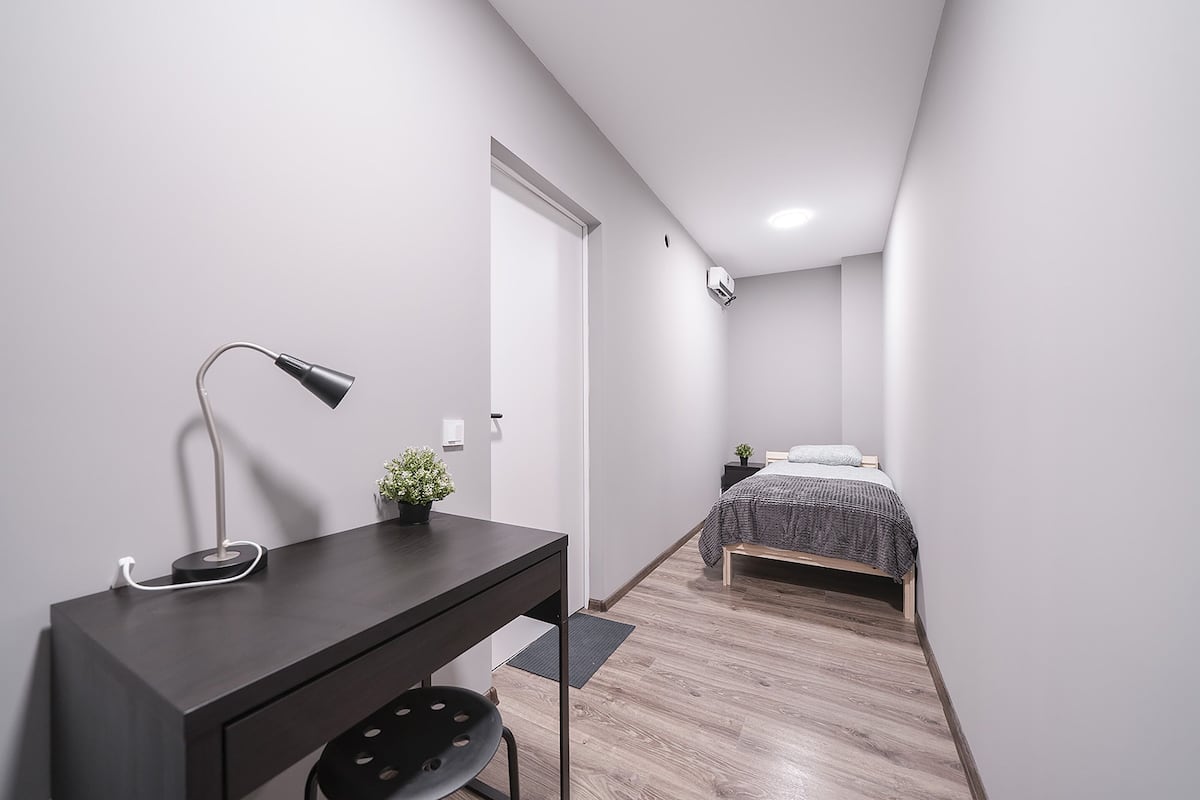 Quarto moderno e estreito, com escrivaninha, cadeira e mesa de cabeceira.