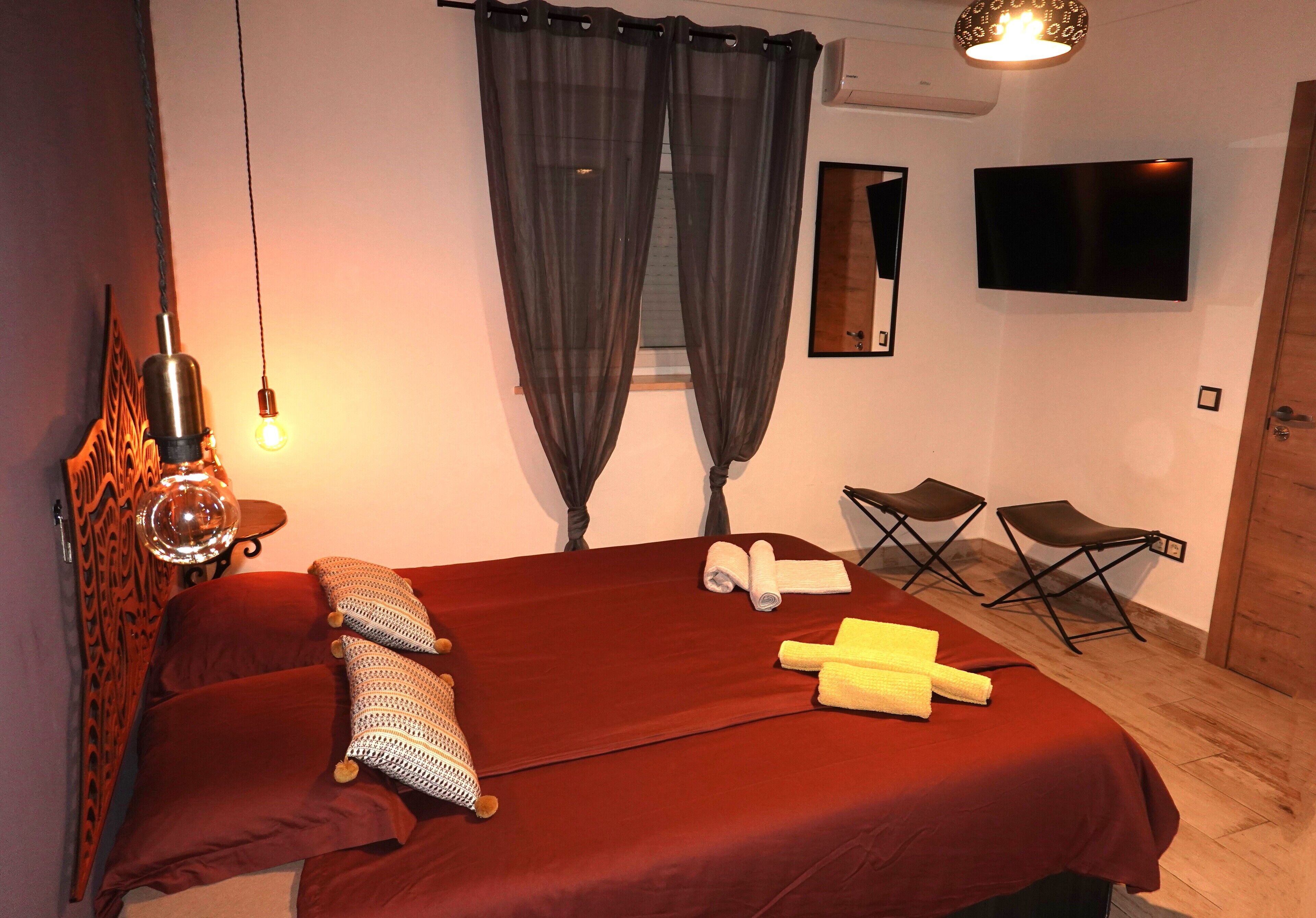 Quarto com uma cama grande, duas cadeiras, uma televisão e uma janela com cortinas leves.