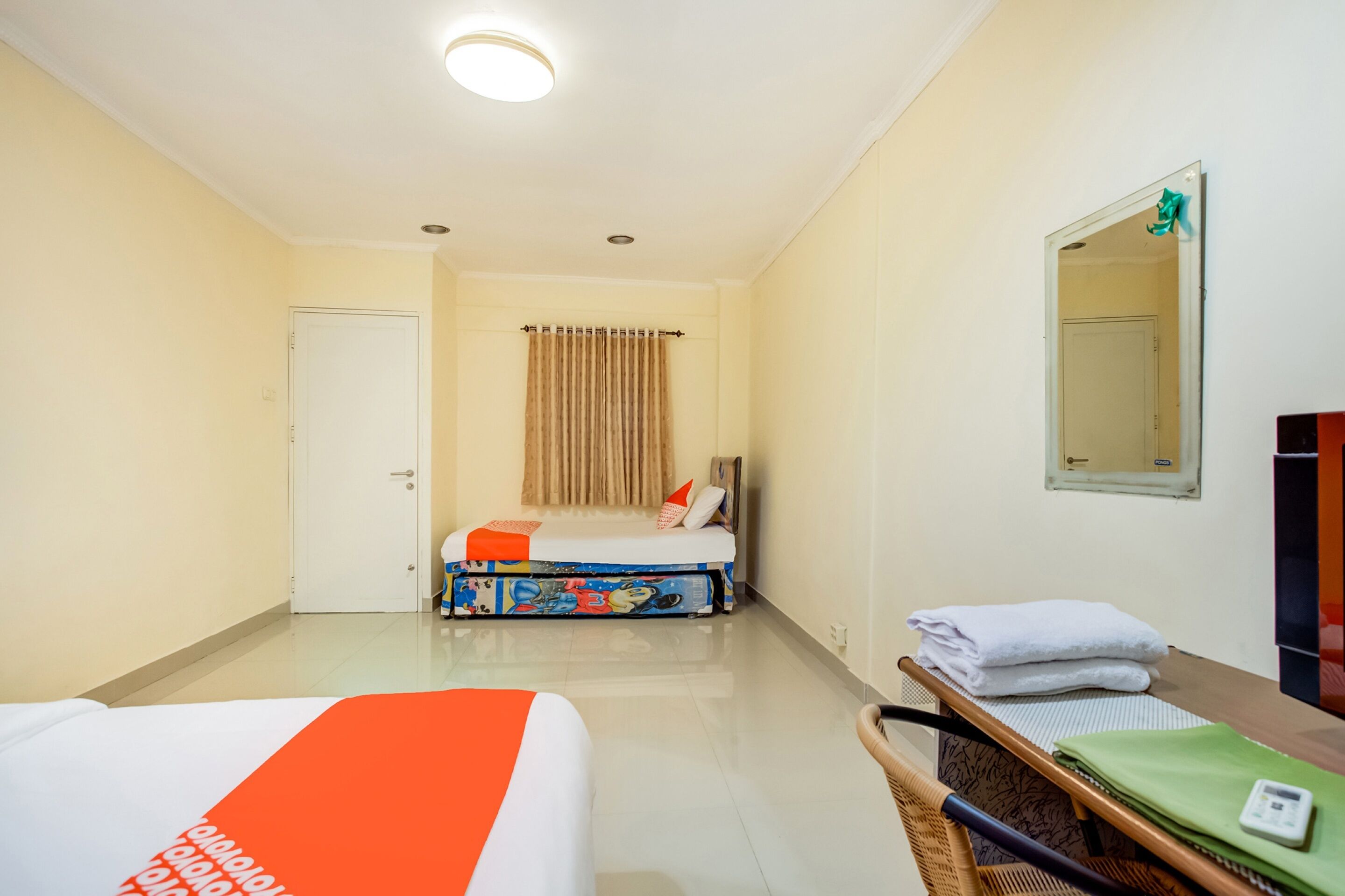 Quarto de hotel com cama, uma cadeira, uma mesa, espelho, porta e cortina.