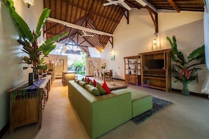 Uma sala de estar ampla com teto alto, sofá verde, móveis de madeira e plantas grandes.