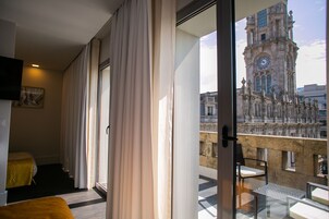Quarto de hotel com varanda, uma cama, uma cadeira e vista para uma torre do relógio histórica.