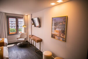 Quarto de hotel com cama, uma cadeira, escrivaninha, janela com cortinas e um quadro na parede, com vista para edifícios.