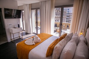 Quarto de hotel com uma cama grande, uma mesa com televisão de tela plana e uma varanda com vista para a cidade.