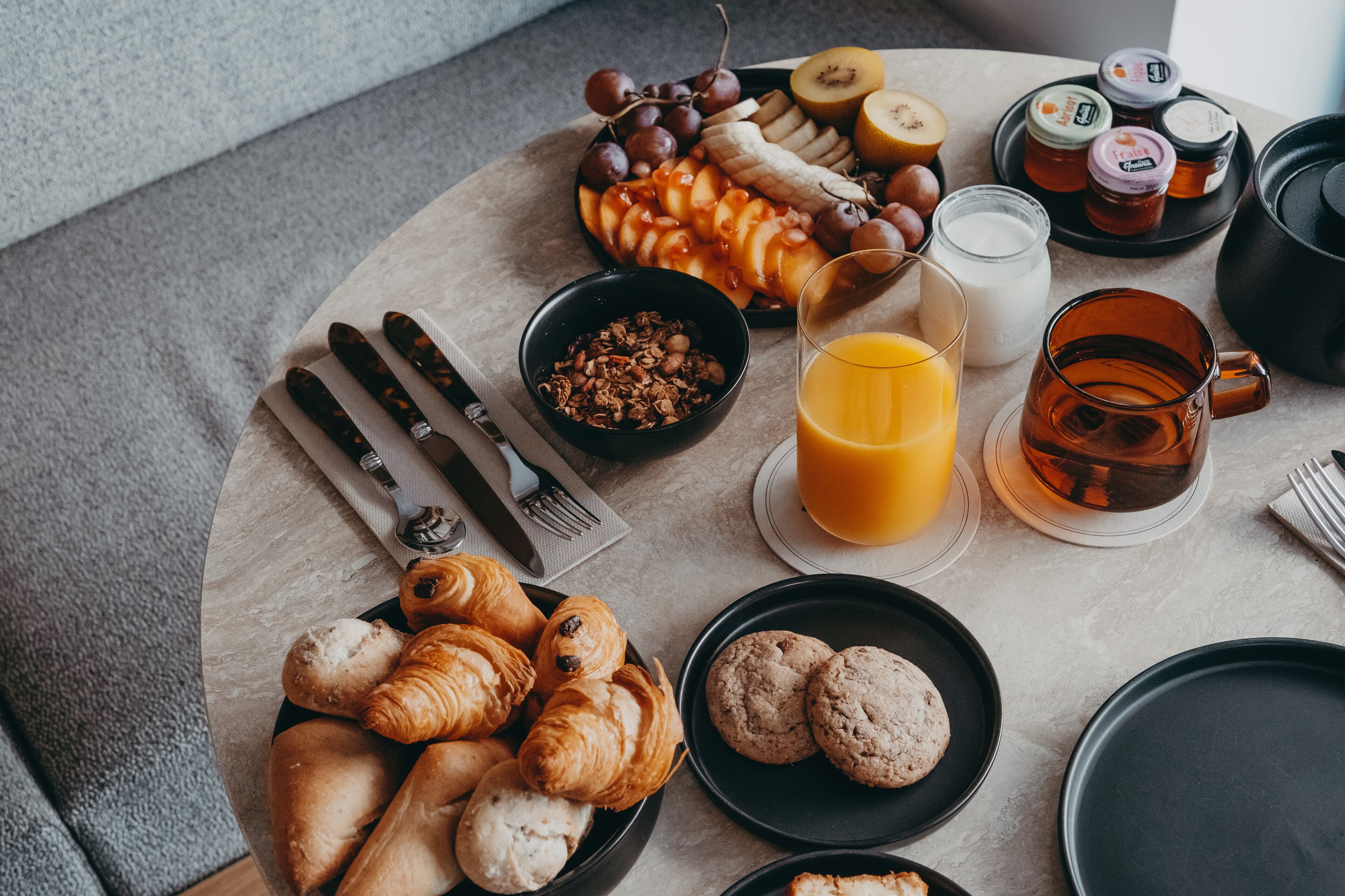 Disposição de café da manhã com croissants, biscoitos, frutas, mel e uma xícara de café em uma mesa redonda.