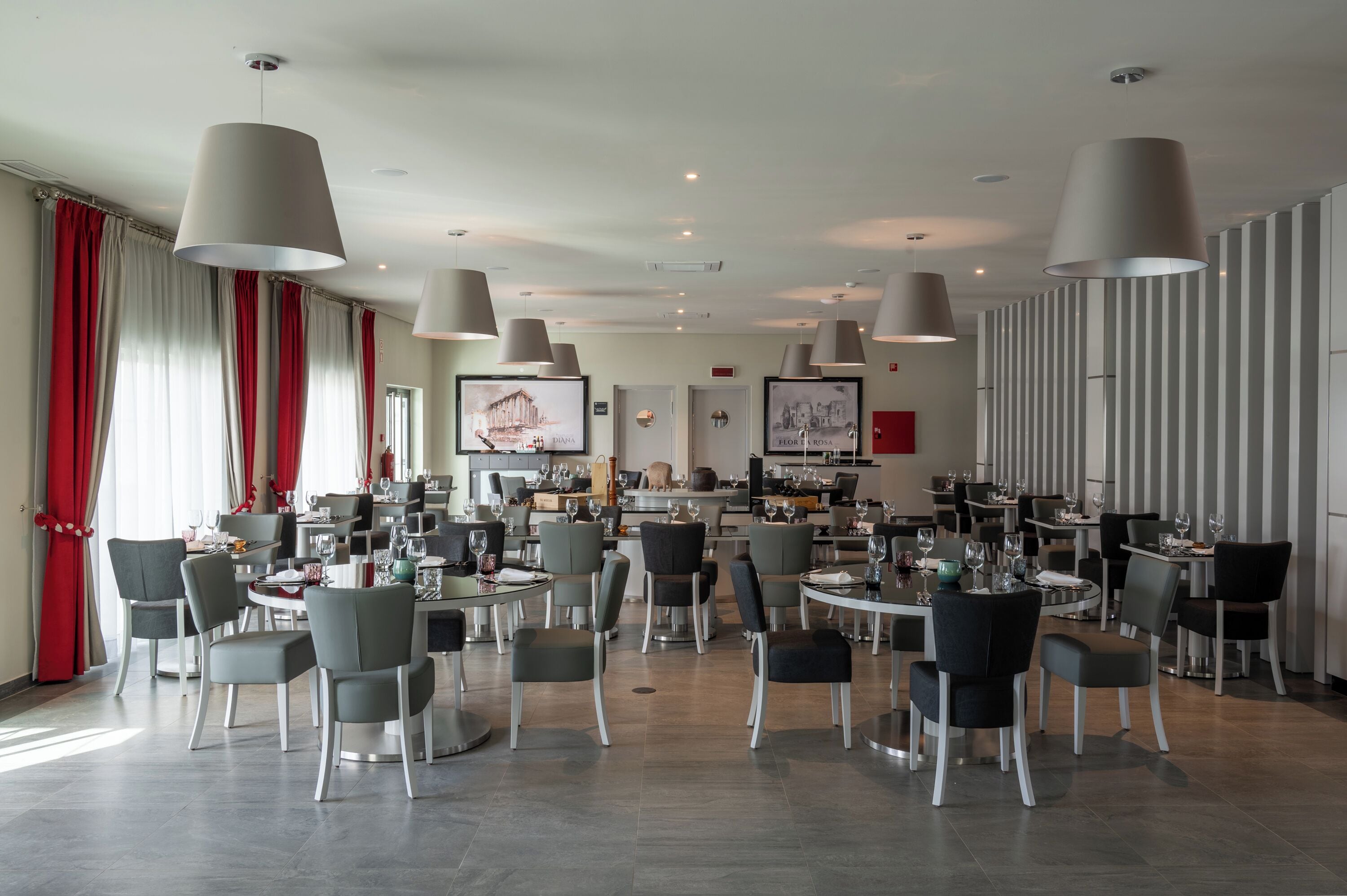 Uma área de jantar moderna com mesas e cadeiras redondas, grandes luminárias pendentes e uma cozinha ao fundo.