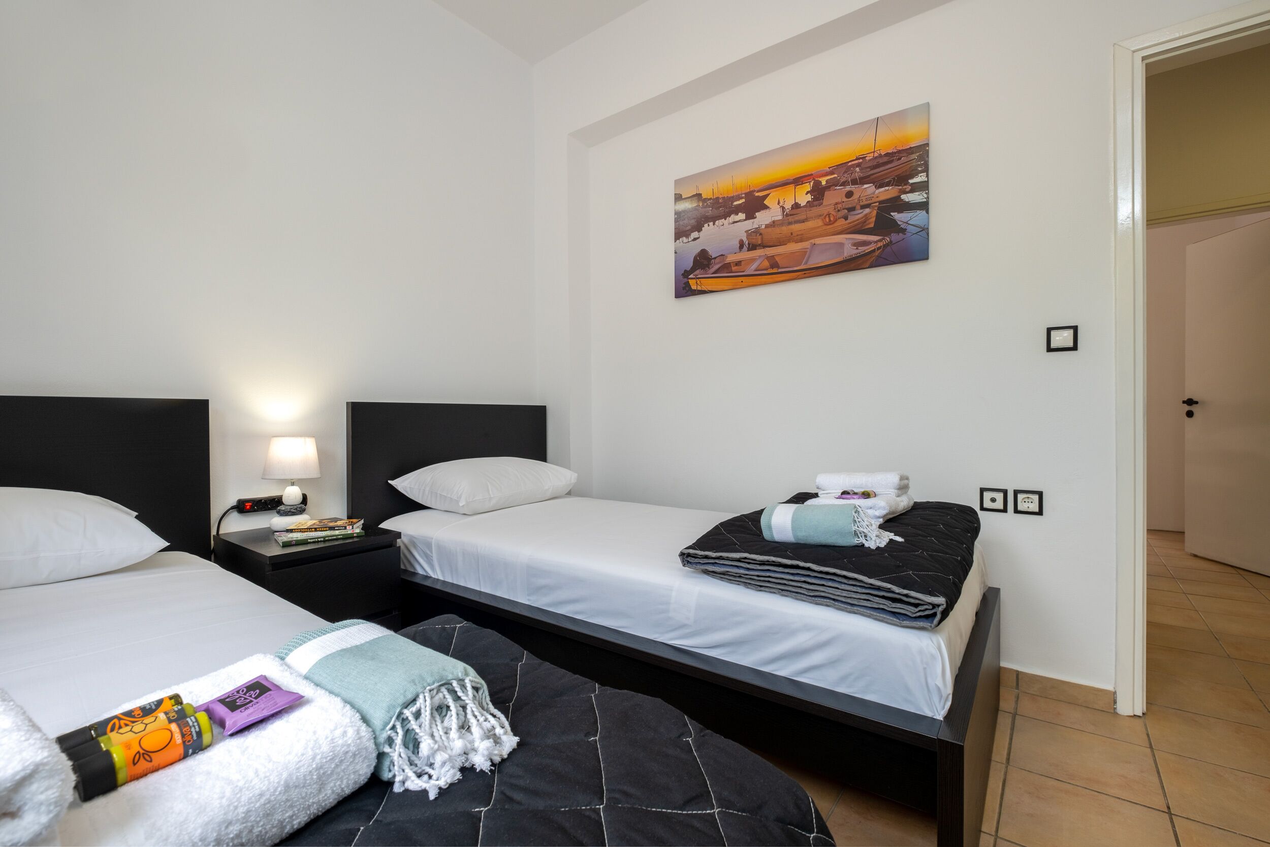 Quarto de hotel com duas camas de solteiro, mesa de cabeceira com abajur, um quadro na parede e uma porta à direita.