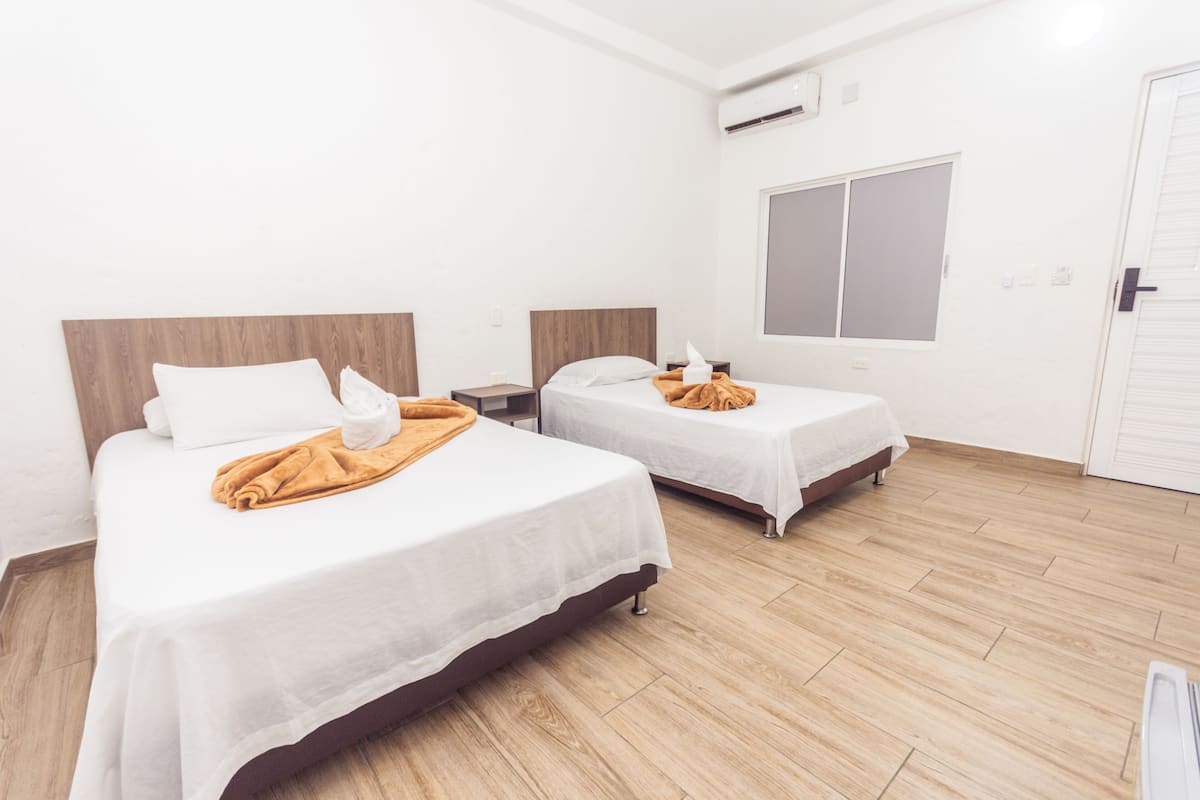 Quarto de hotel com duas camas, cada uma com colcha branca e manta marrom. Há uma janela com persianas, uma porta branca e uma pequena mesa de cabeceira com uma lâmpada.