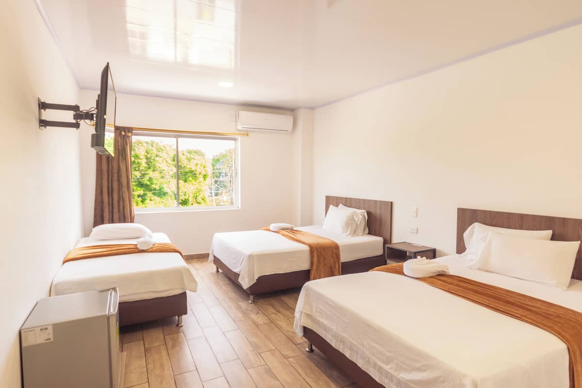 Quarto de hotel com três camas, televisão, ar condicionado e janela com vista para áreas verdes.
