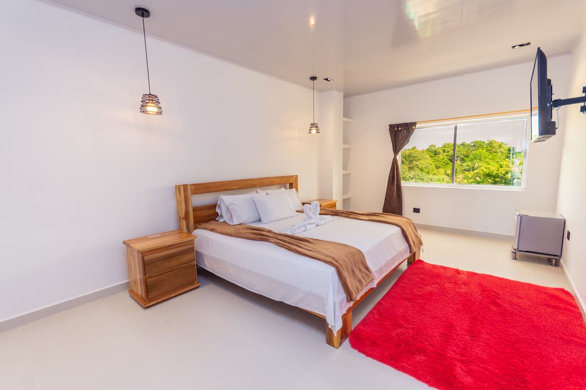Quarto moderno com cama de madeira, roupa de cama branca, tapete vermelho e janela com vista para o verde.
