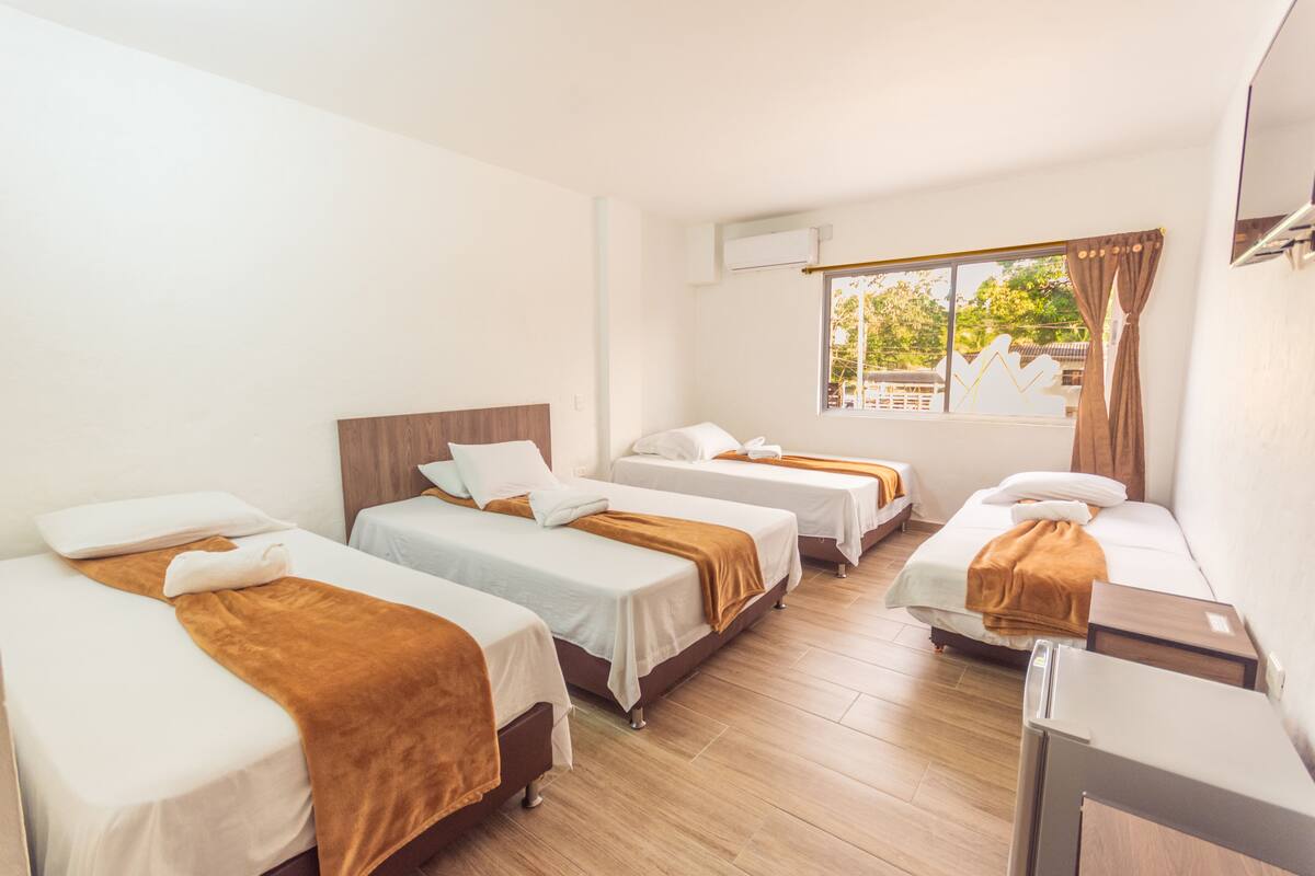 Quarto de hotel com quatro camas de solteiro, piso de madeira, janela com cortinas e uma pequena escrivaninha.