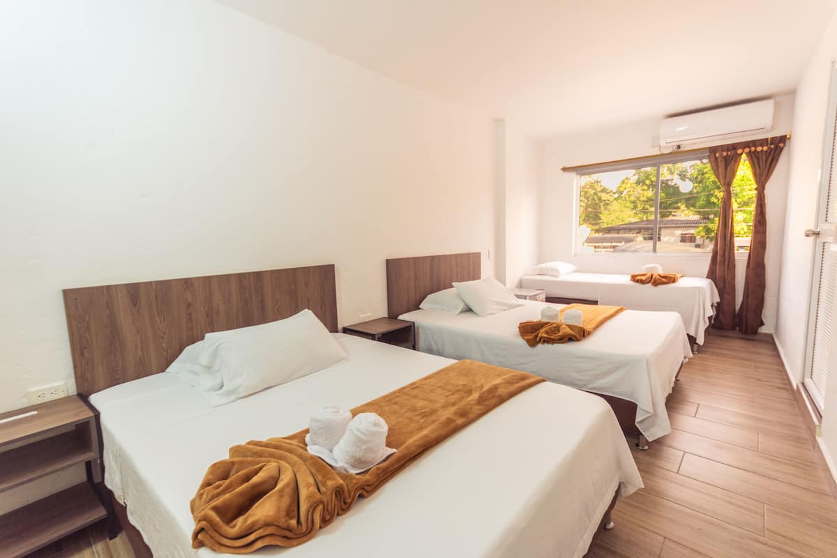 Quarto de hotel com três camas, uma janela com cortinas e um aparelho de ar condicionado.