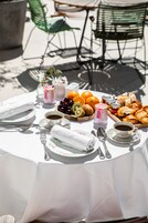 Mesa posta com itens de café da manhã, incluindo frutas, doces e bebidas, cercada por cadeiras e mesa ao ar livre.