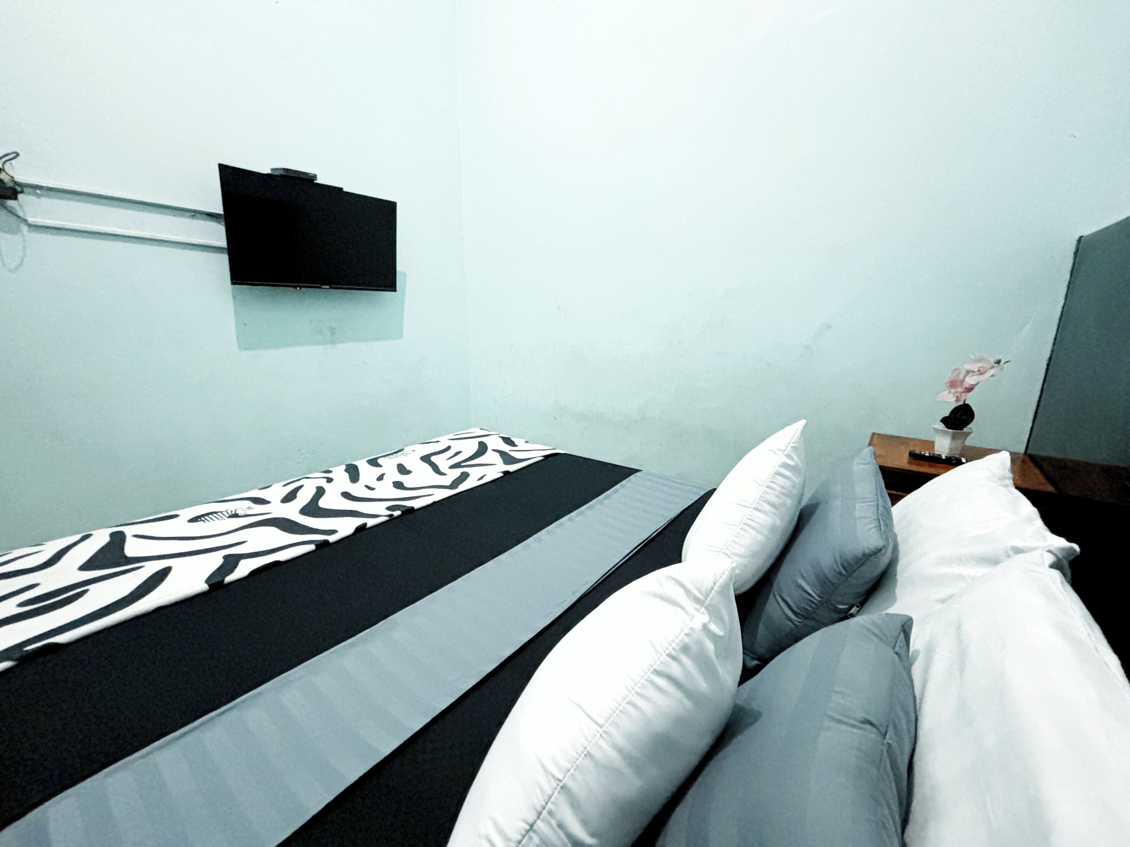 Quarto com cama, almofadas, mesa de cabeceira com um vaso e uma televisão na parede.