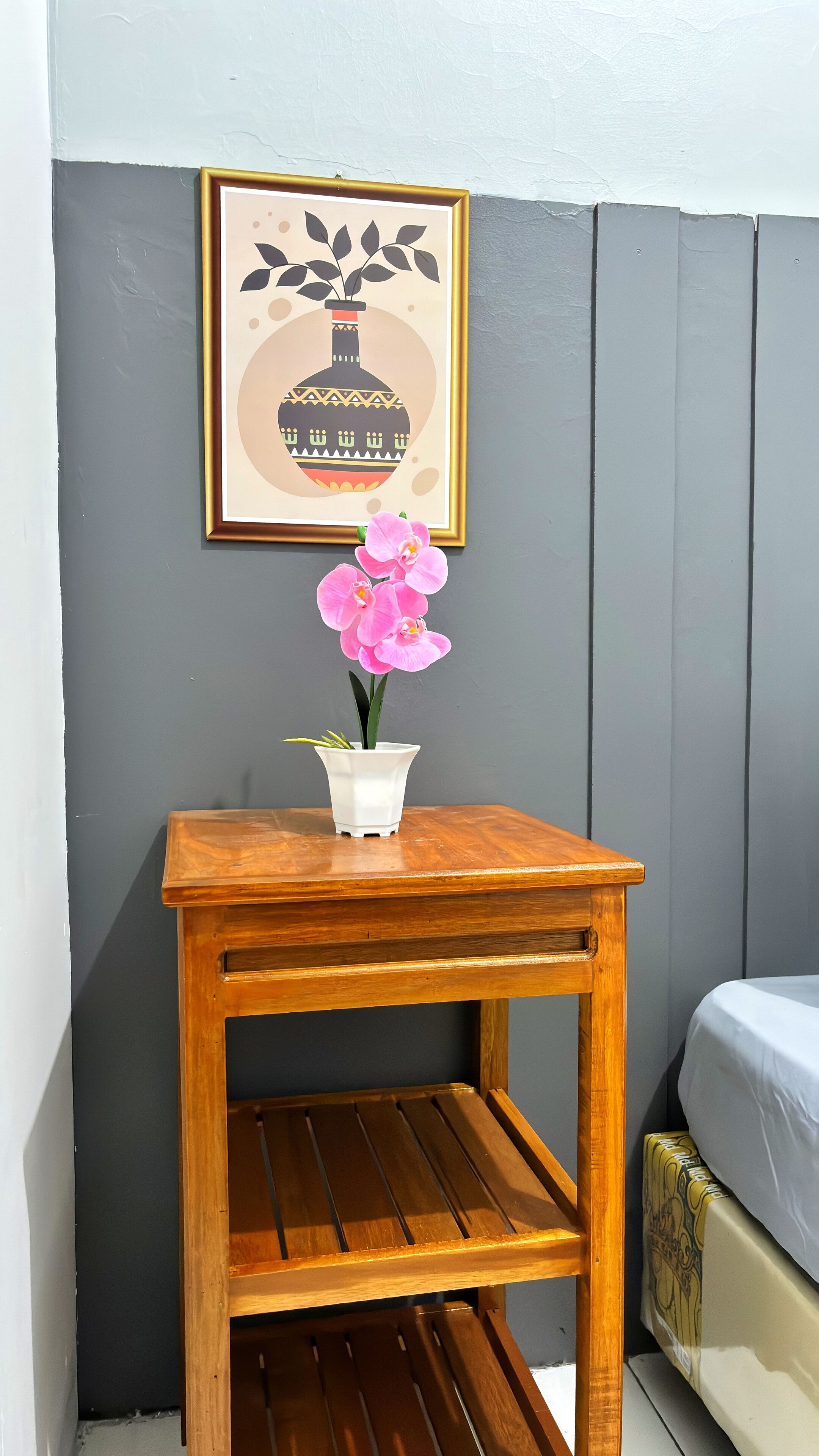 Uma mesinha de cabeceira de madeira com um vaso branco contendo flores cor-de-rosa, posicionada em frente a uma parede cinza com um quadro emoldurado acima.