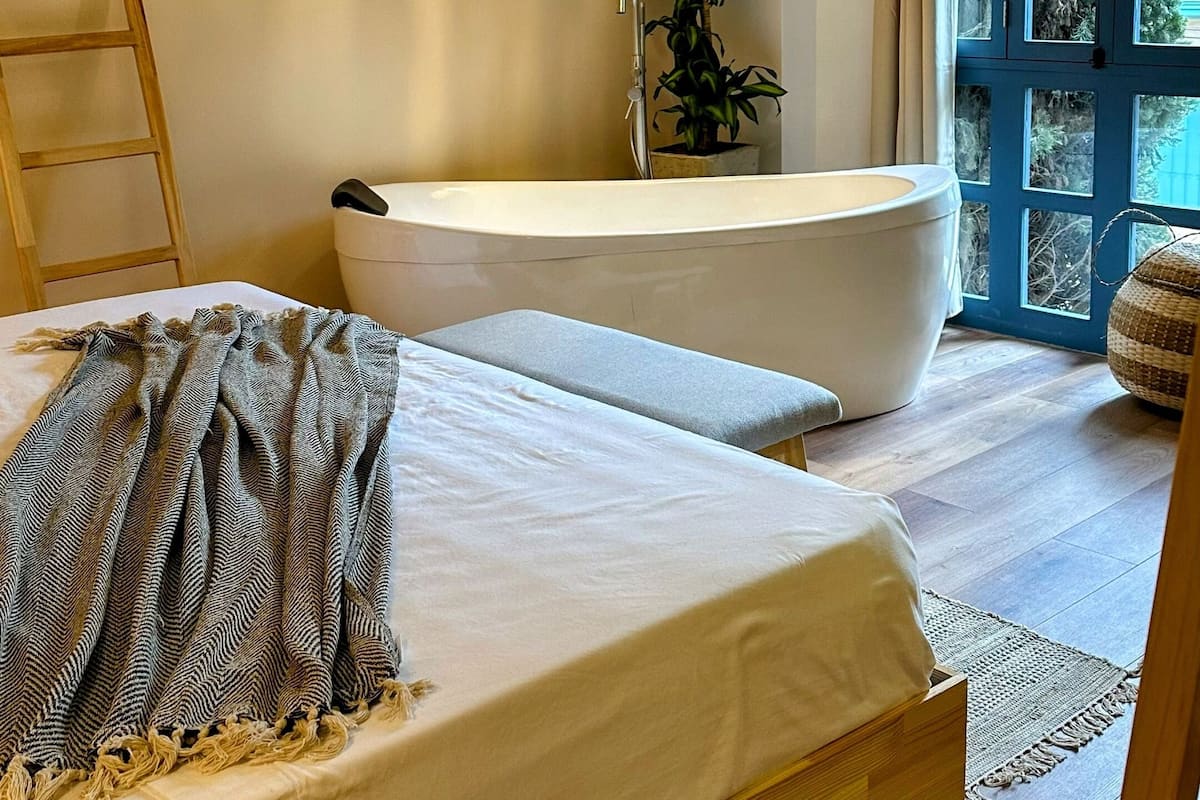 Quarto com cama grande, banheira, luminária pendente e vista pela janela.