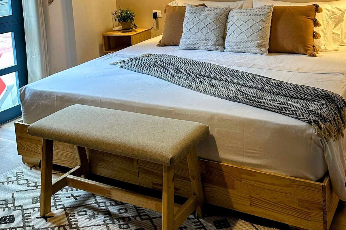 Quarto com cama grande, um banquinho, tapete estampado, luminária de macramé na parede e luminária pendente.