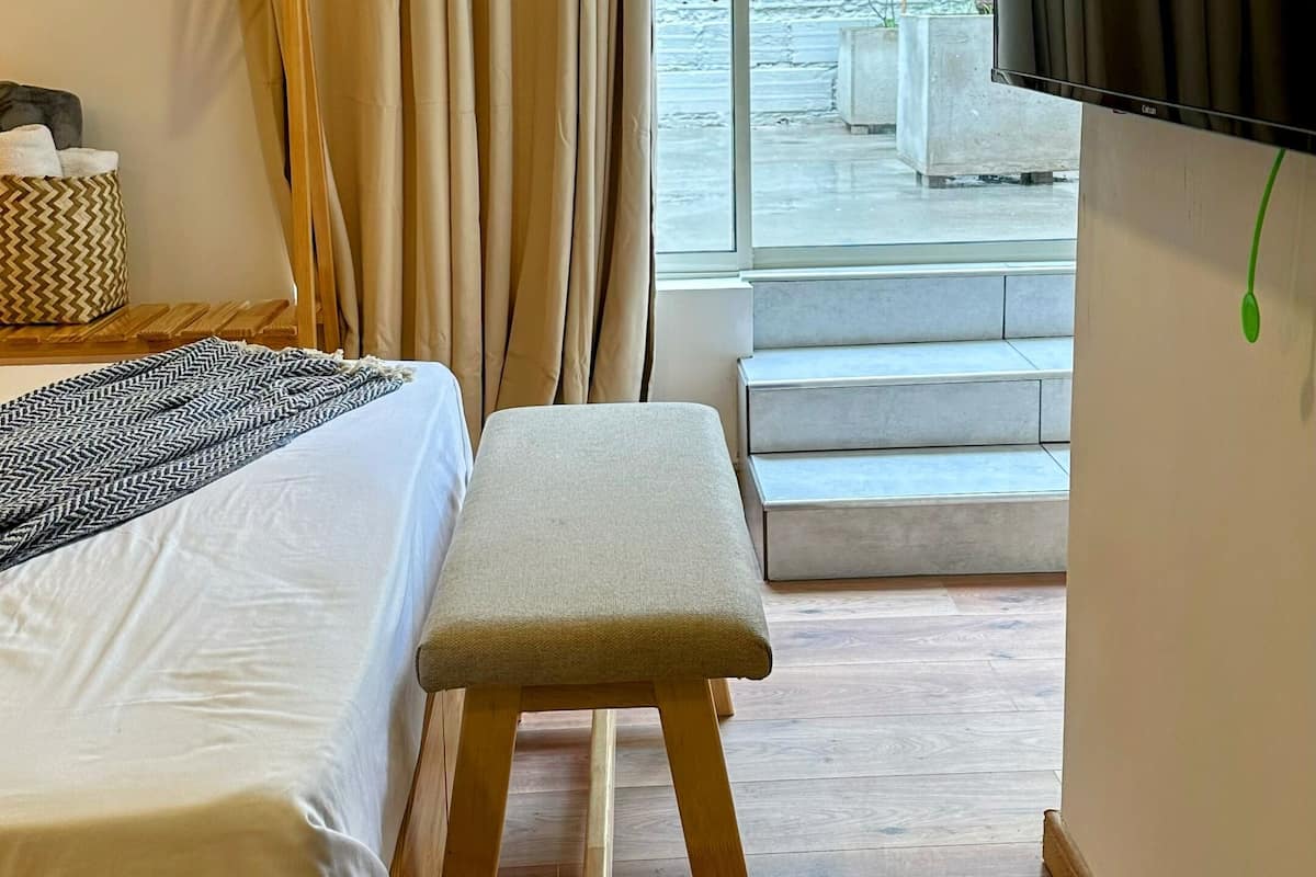 Quarto moderno com cama de madeira, um banco, uma televisão e uma janela grande com cortinas.