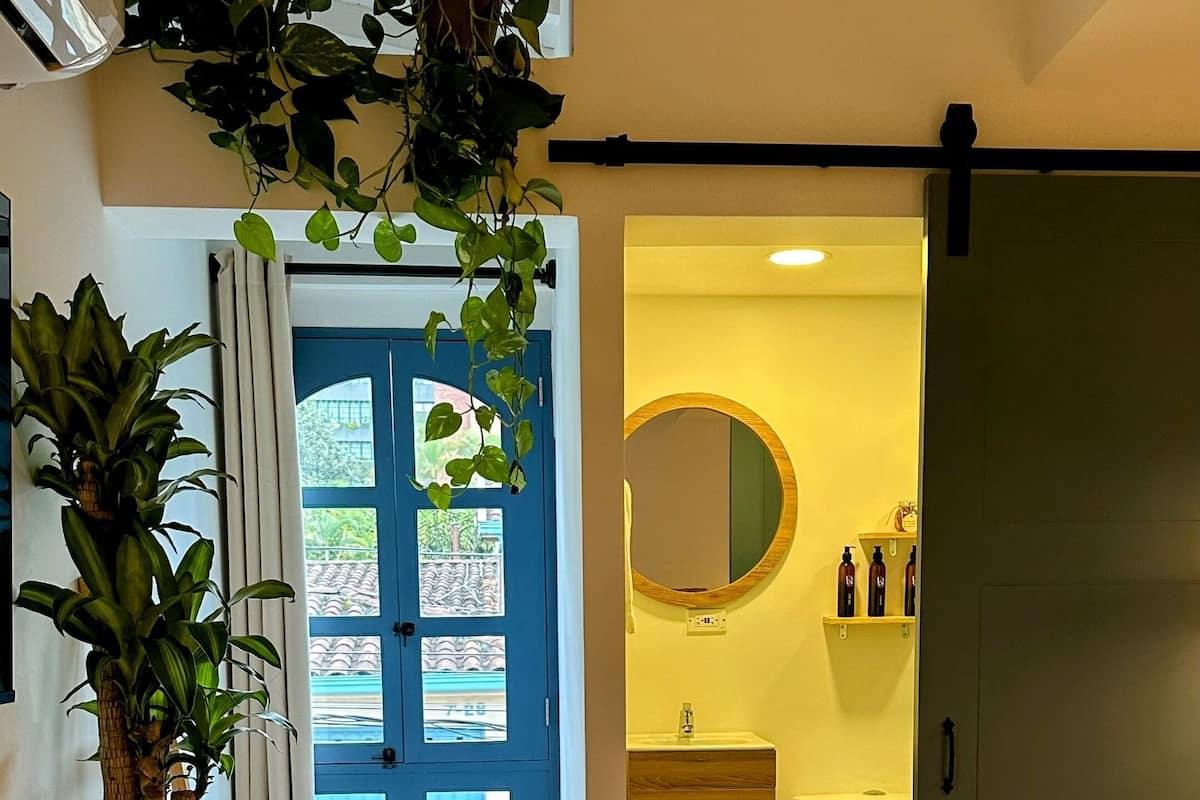 Quarto moderno com cama, uma cadeira, uma planta em vaso grande e vista do banheiro por uma porta aberta.