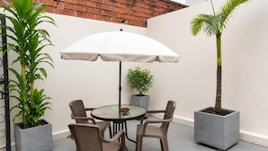 Um pátio com uma mesa redonda, quatro cadeiras, um guarda-sol branco e plantas em vasos.