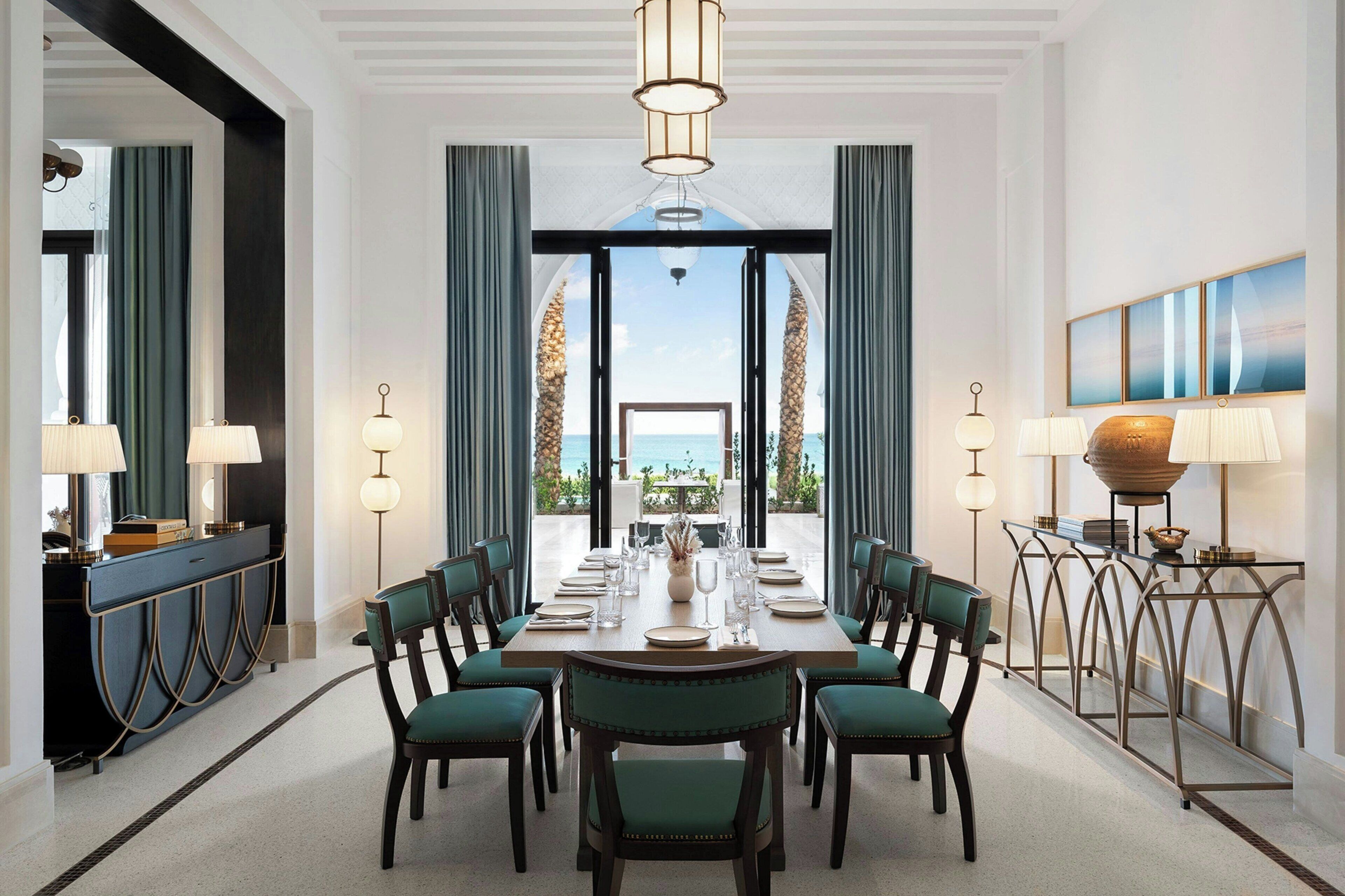 Uma área de refeições com uma mesa comprida posta para seis pessoas, ladeada por cadeiras, e vista para o mar através de grandes janelas.