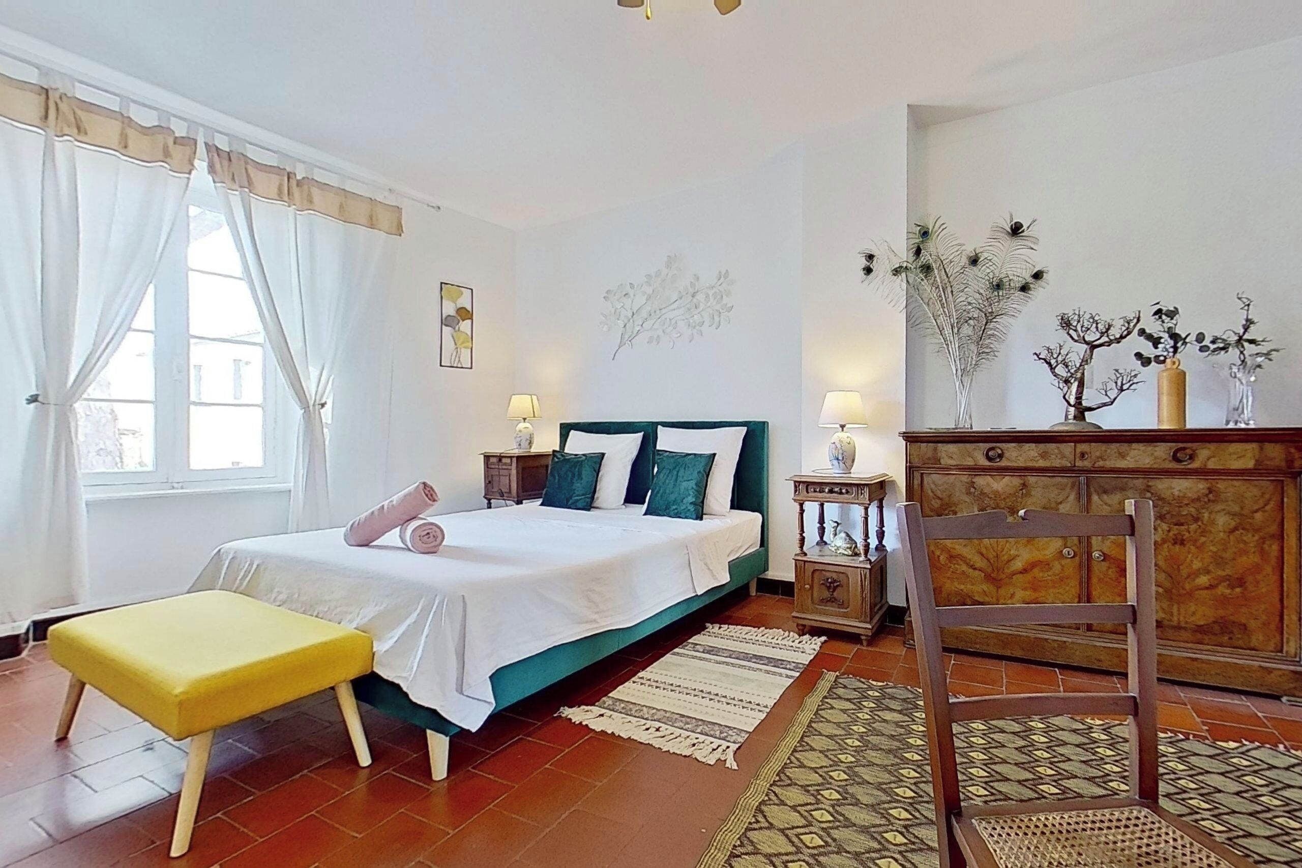 Quarto com cama, mesas de cabeceira, uma cadeira, janela com cortinas e arte decorativa na parede.