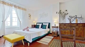 Quarto com cama, mesas de cabeceira, uma cadeira, janela com cortinas e arte decorativa na parede.