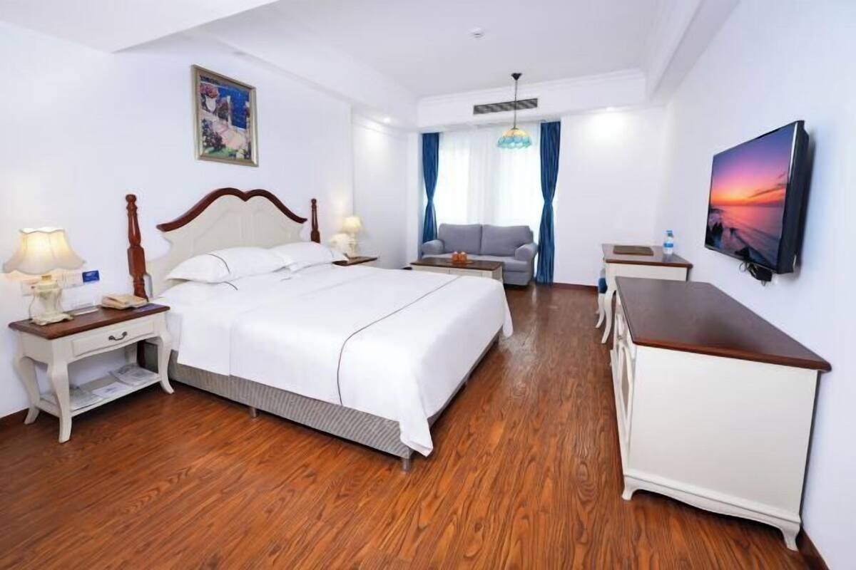 Quarto de hotel com cama, mesas de cabeceira, escrivaninha, poltrona, televisão e janela com cortinas.