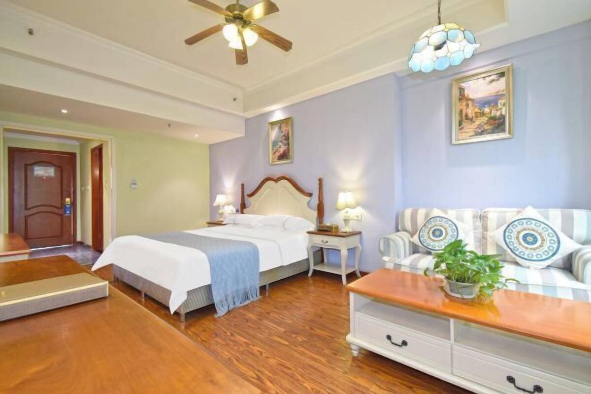 Quarto de hotel com cama, mesas de cabeceira, sofá, mesa de centro de madeira e ventilador de teto.