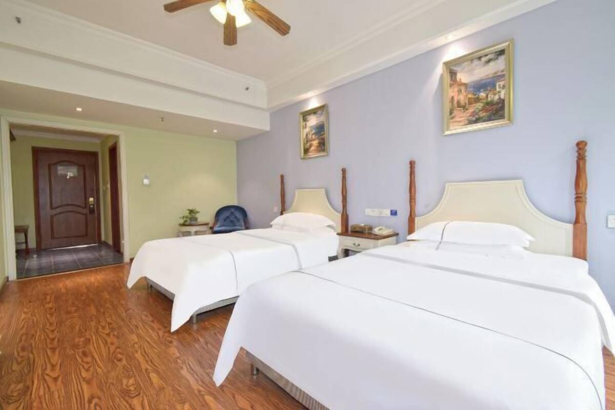 Quarto de hotel com duas camas, piso de madeira, ventilador de teto e porta para o corredor.