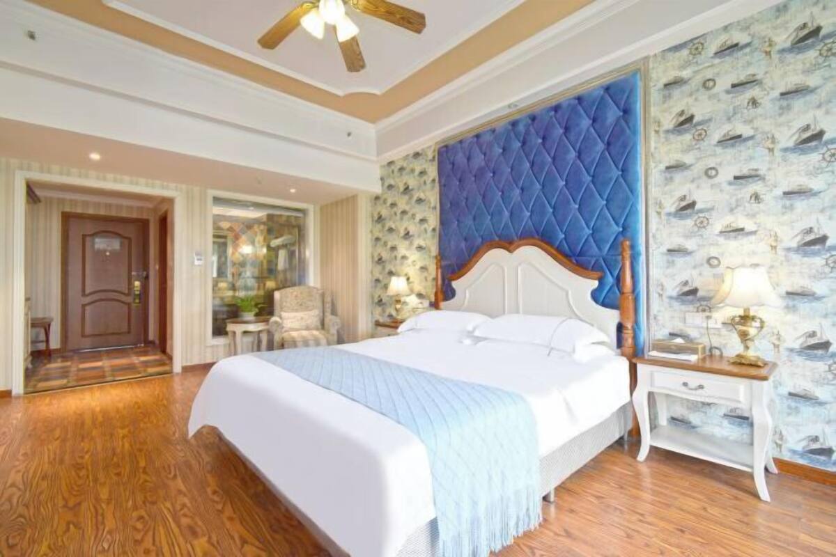 Quarto de hotel com uma cama grande, cabeceira estofada, piso de madeira, uma mesinha com abajur e papel de parede estampado.