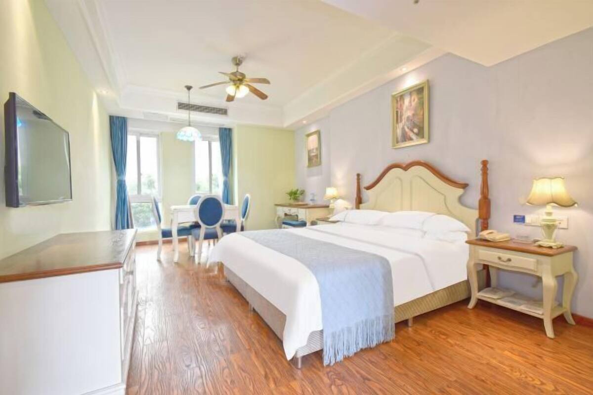 Quarto de hotel com cama grande, escrivaninha, cadeira, ventoinha no teto e televisão na parede.
