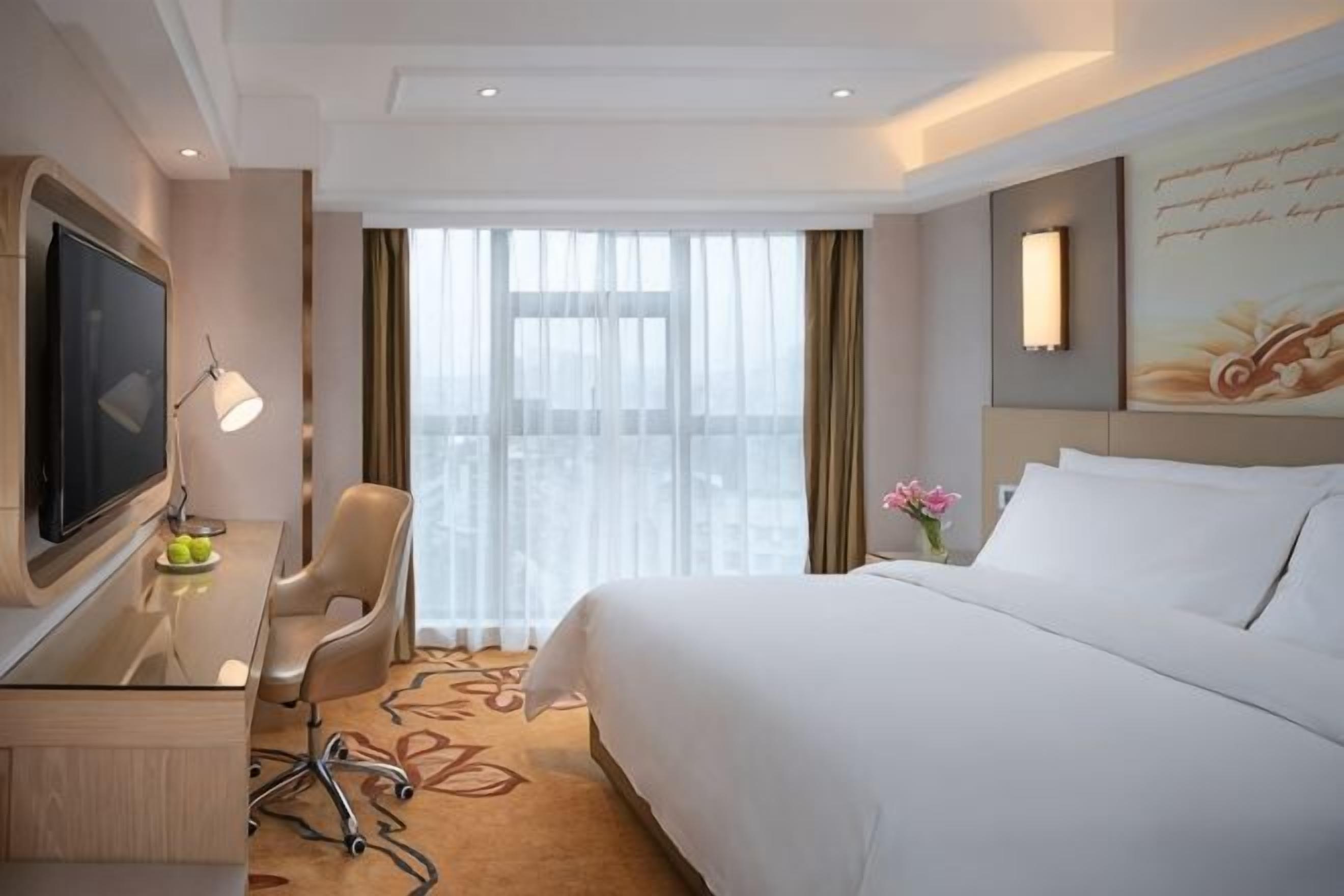 Quarto de hotel com cama, escrivaninha com televisão, cadeira, luminária de mesa, vaso de flores e janela com cortinas.