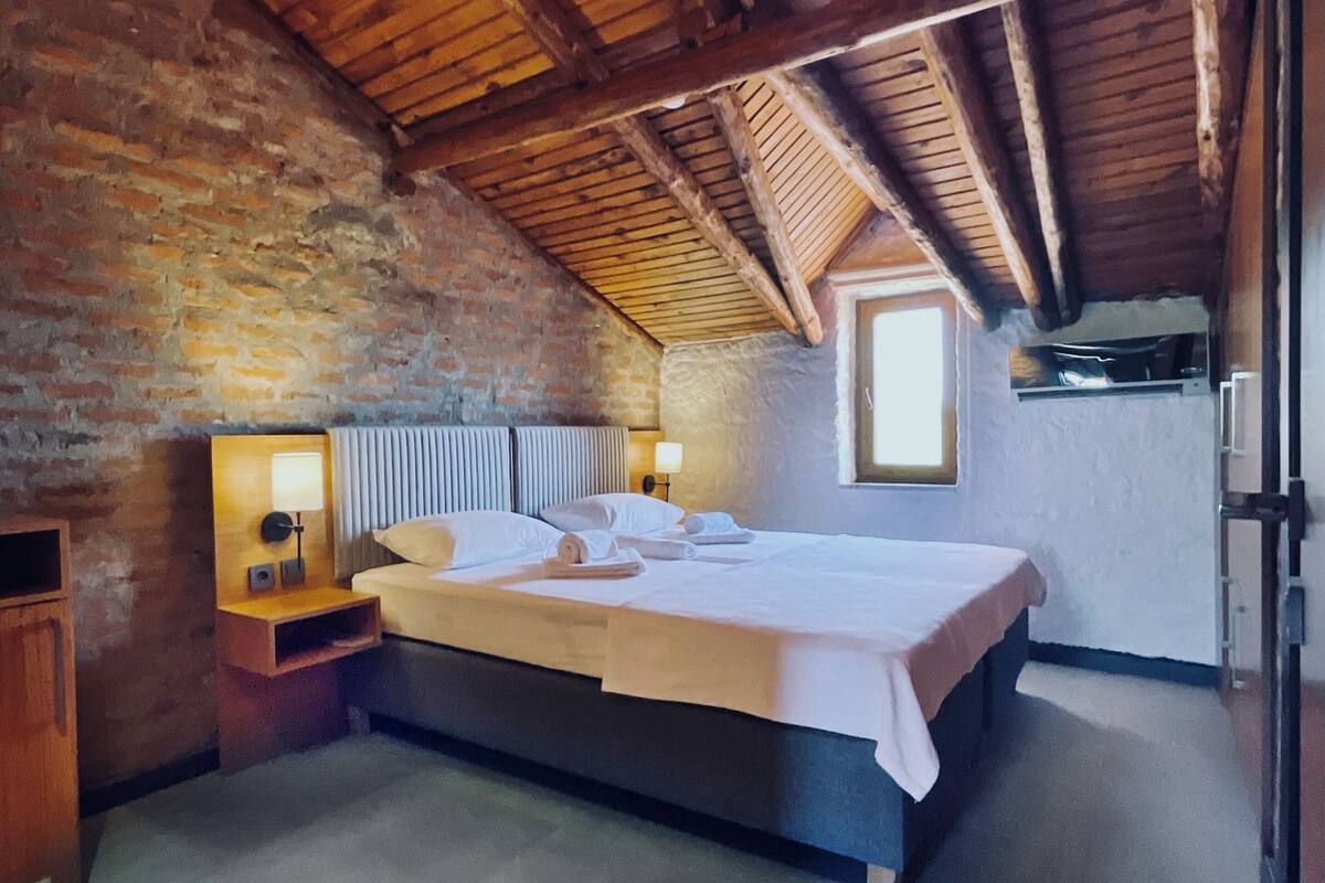 Quarto com teto de madeira, parede de tijolo, cama com roupa de cama branca, mesa de cabeceira com abajur e janela.