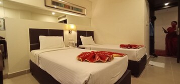 Quarto de hotel com duas camas, cada uma com colcha branca e manta vermelha. Tem uma mesinha de cabeceira com luminária, telefone e uma pequena mesa. O quarto tem um design moderno, com um espelho grande acima das camas e uma porta que dá para outro espaço.