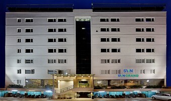 Hotel com vários andares, entrada central, varandas e área de estacionamento com motociclos.