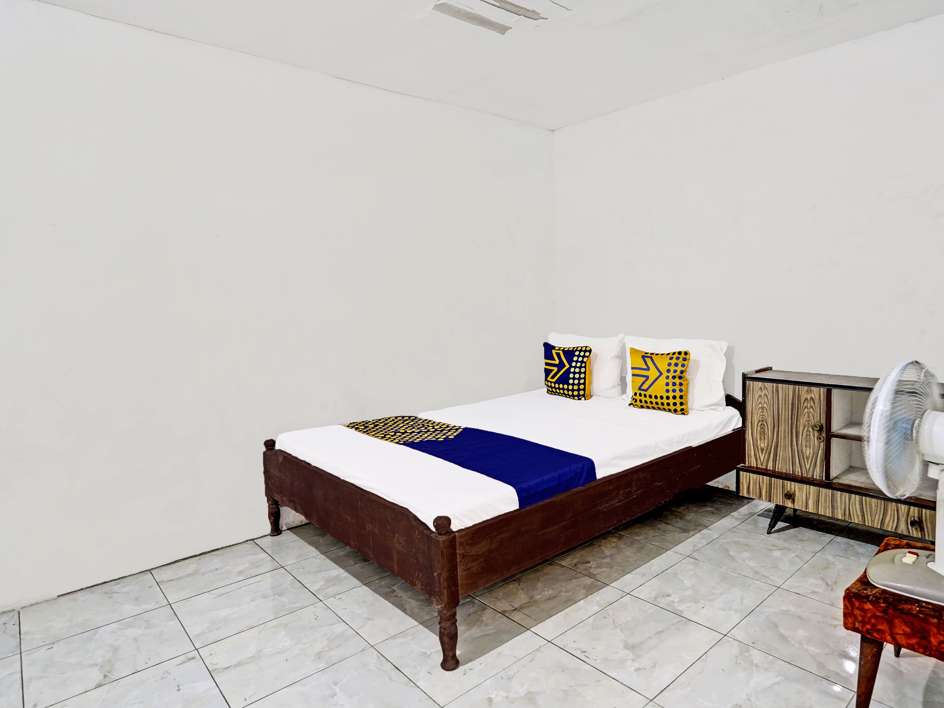 Uma cama de solteiro com estrutura de madeira, roupa de cama branca e almofadas estampadas. Um pequeno armário de madeira e um ventilador branco estão visíveis no quarto.