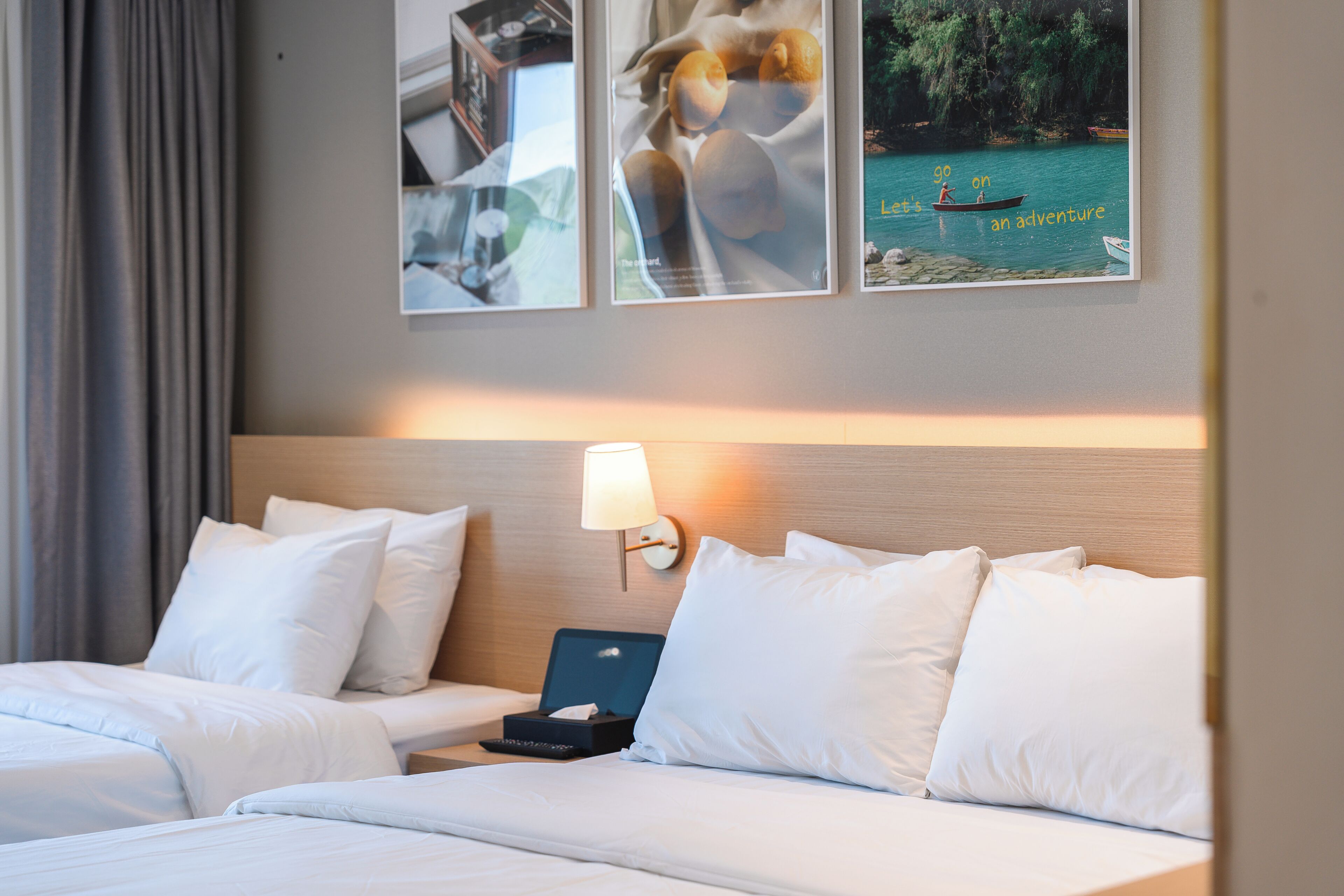 Quarto de hotel com duas camas, uma mesinha de cabeceira com abajur e três quadros na parede.