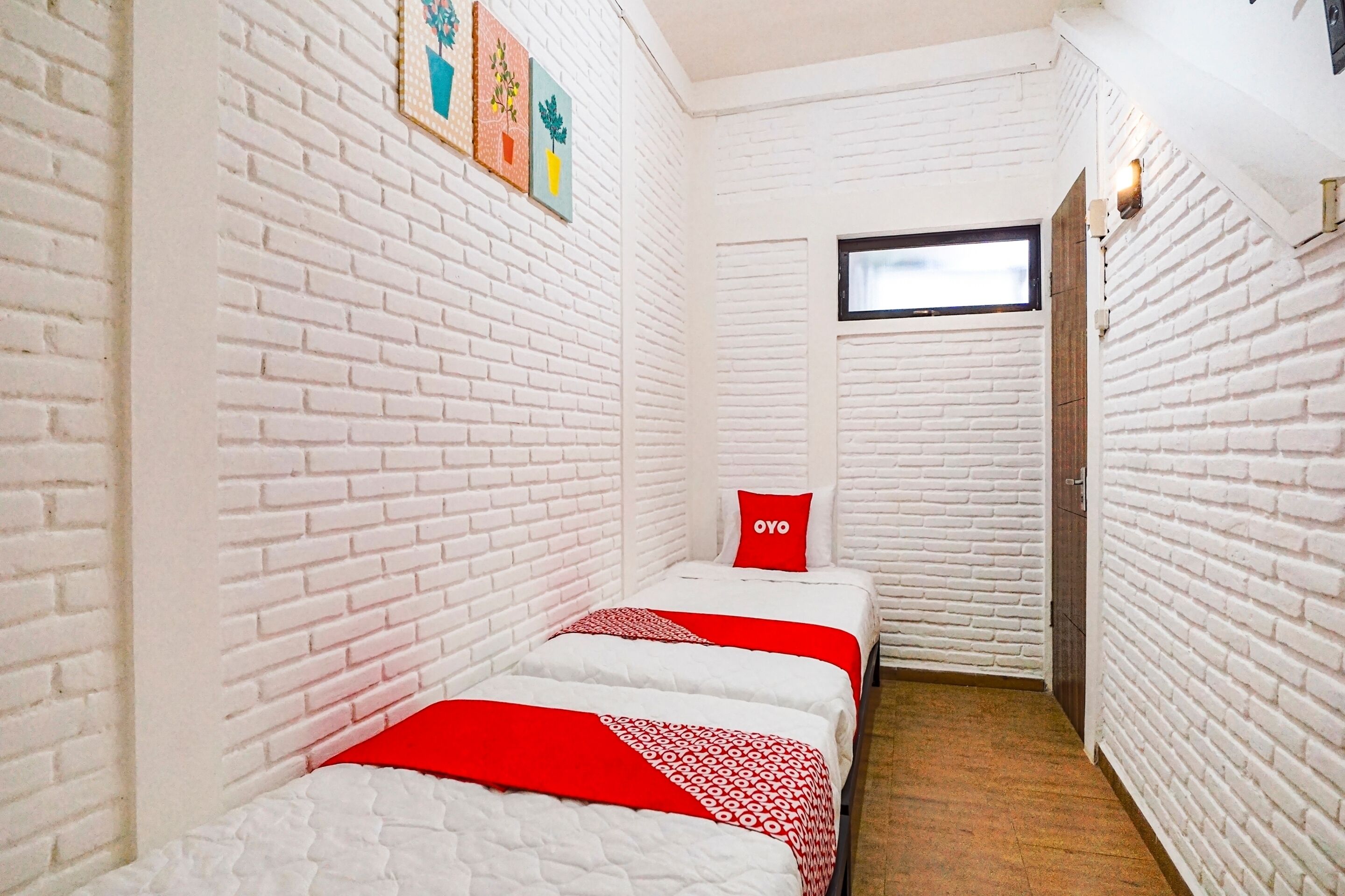 Um quarto pequeno com paredes de tijolo branco, duas camas com roupa de cama vermelha e branca, e uma janela.