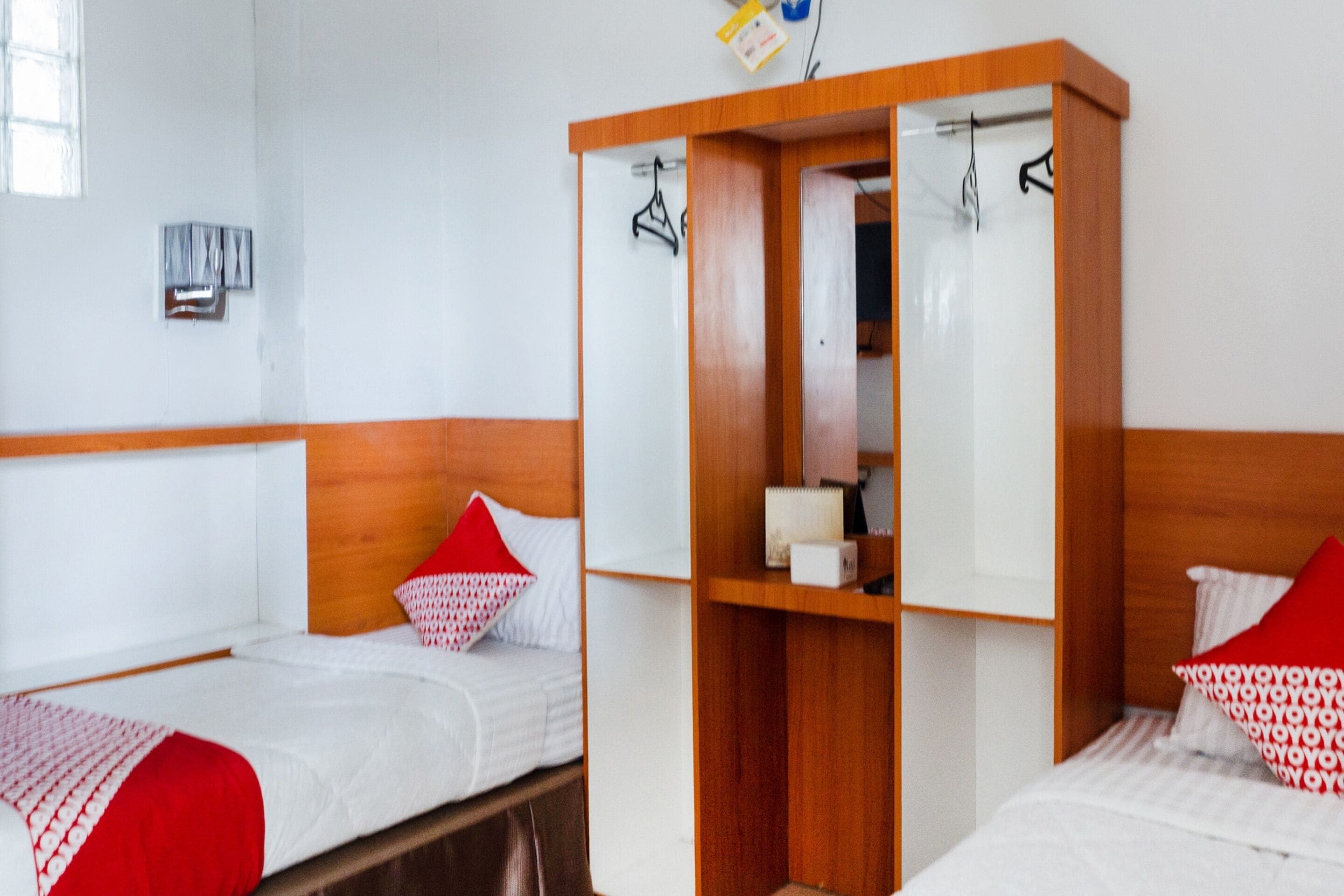 Quarto de hotel com duas camas de solteiro, cada uma com almofada estampada em vermelho e branco, e um guarda-roupa de madeira com espelho.